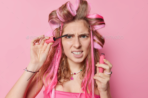 Irritated woman with long hair does hairstyle applies mascara on her ...
