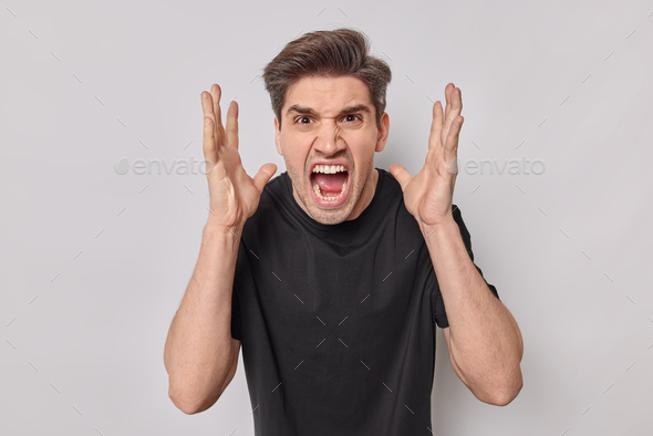 Outraged young man yells angrily keeps mouth opened raises hands being ...