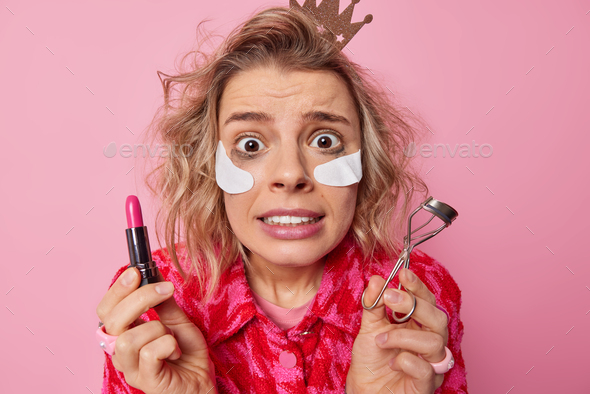 Worried anxious young woman holds lipstick and eyelashes curler for ...