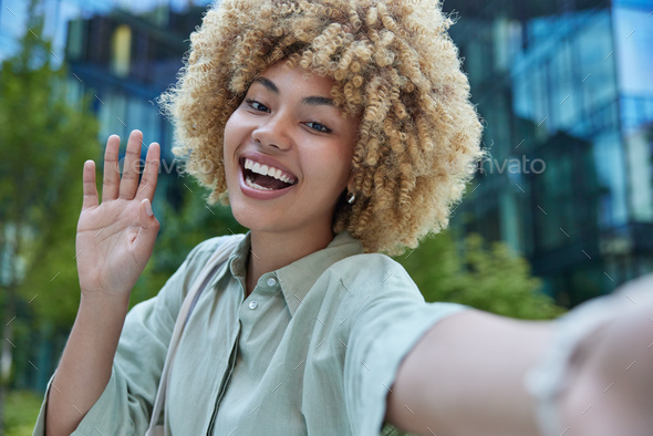 Self portrait of cheerful curly haired woman waves hello keeps arm ...