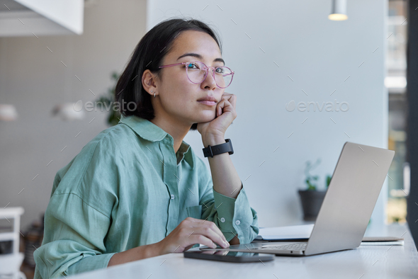 Thoughtful Asian woman works freelancer uses laptop computer smartphone ...