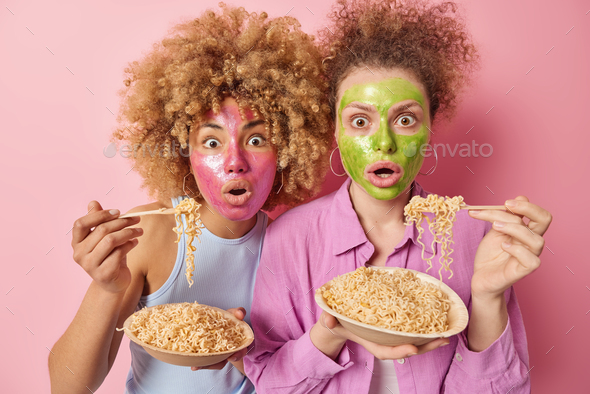 Impressed curly haired woman eat pasta apply beauty masks on face stand ...
