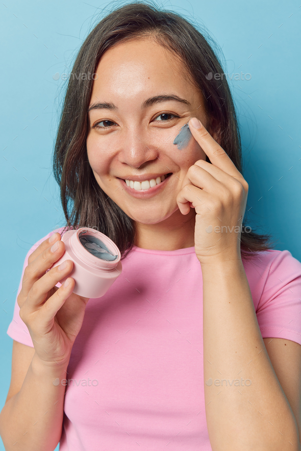 Happy dark haired Asian woman applies nourishing cream on face ...