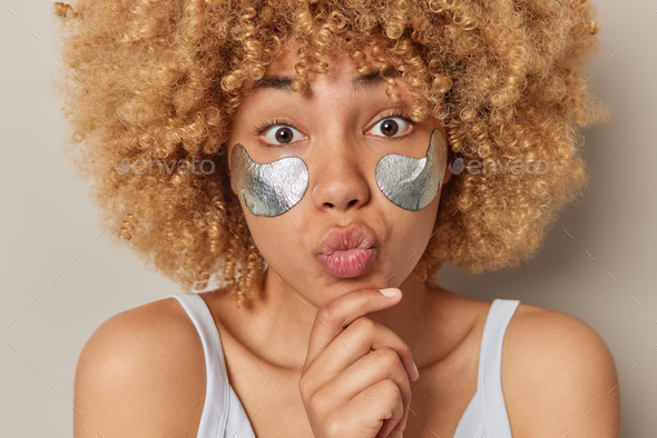 Close up portrait of surprised curly haired woman pouts lips holds chin ...