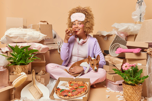 Positive curly haired beautiful woman dressed in domestic clothes eats ...