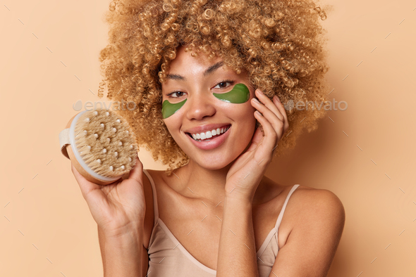 Photo of happy curly haired woman keeps hand on face applies beauty ...