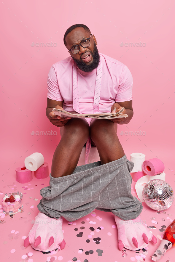 Displeased bearded dark skinned man sits on toilet bowl reads magazine ...