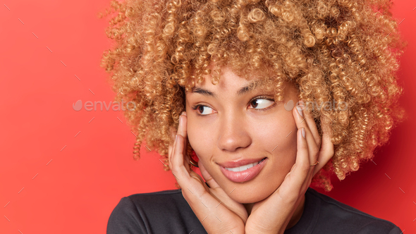 Headshot of beautiful curly woman keeps hands on cheeks concentrated ...