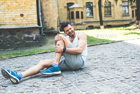handsome sportsman screaming while sitting on pavement and touching ...