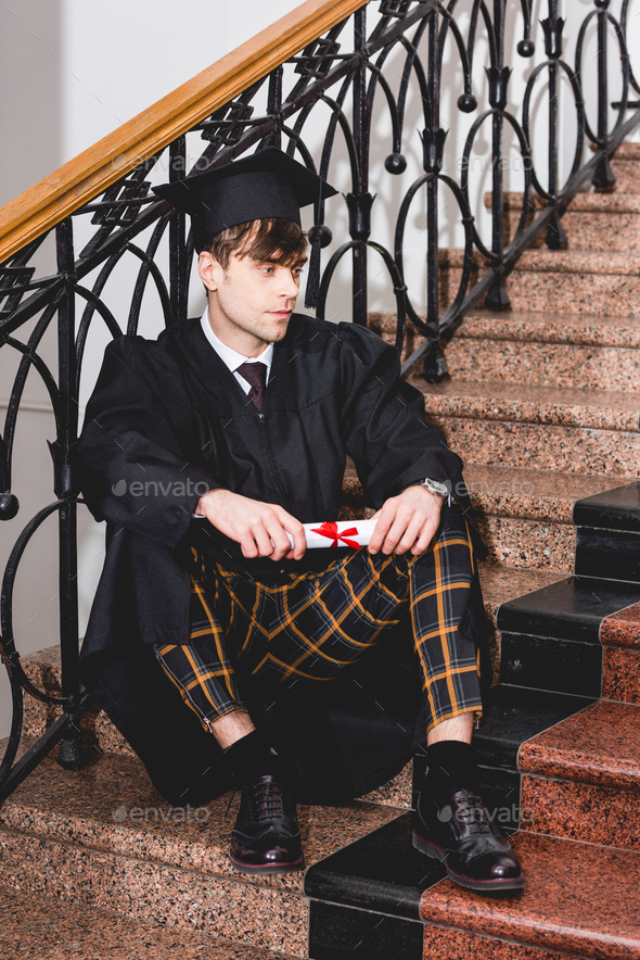 handsome student in graduation gown sitting on stairs and holding ...