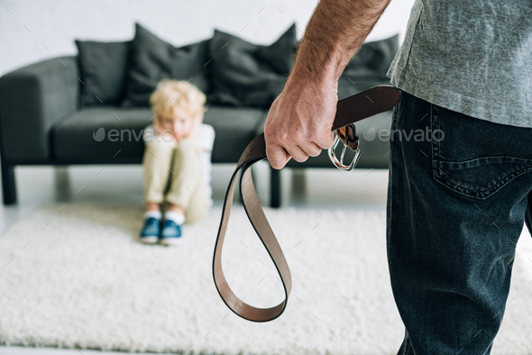 partial view of abusive father holding belt and sad son sitting on ...