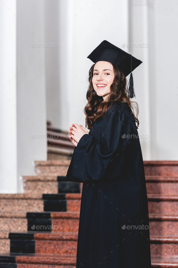 cheerful young woman in graduation cap smiling while standing in ...