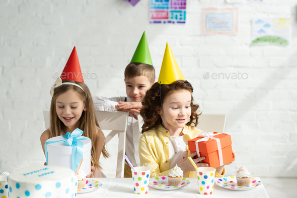 adorable kids in party caps sitting at table with gift boxes during ...