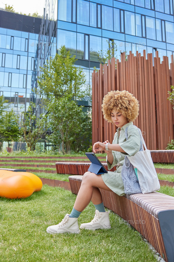Outdoor shot of curly beautiful millennial girl checks time on ...