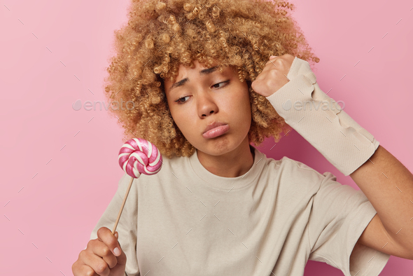 Upset curly haired young woman looks sadly at sweet lollipop on stick ...