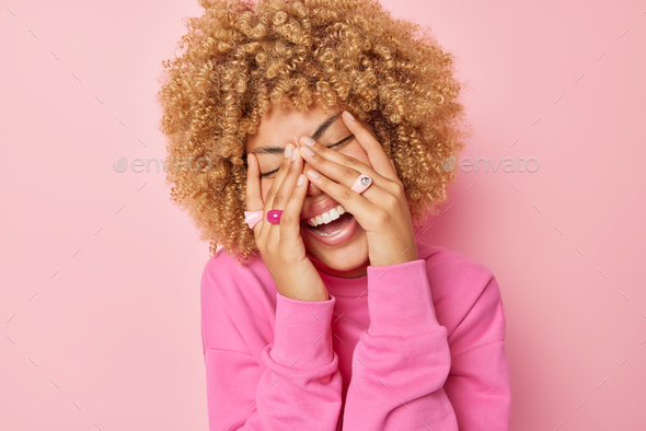 Overjoyed woman with curly hair covers face with hands laughs happily ...