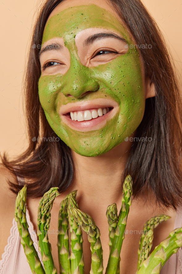 Headshot of brunette young Asian woman applies green facial mask holds ...