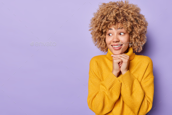 Pretty curious curly woman smiles broadly keeps hands under chin feels ...