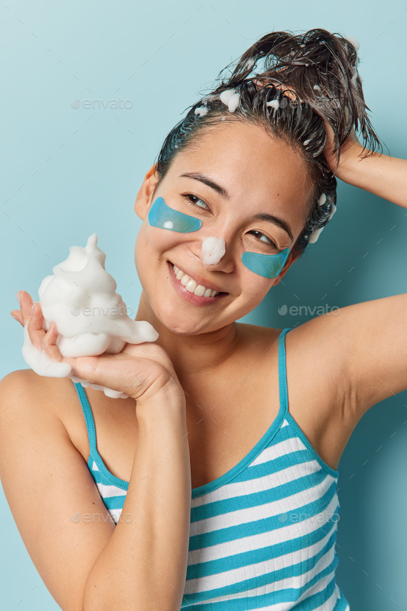 Positive dreamy Asian woman cleans hair with foamy shampoo takes shower ...