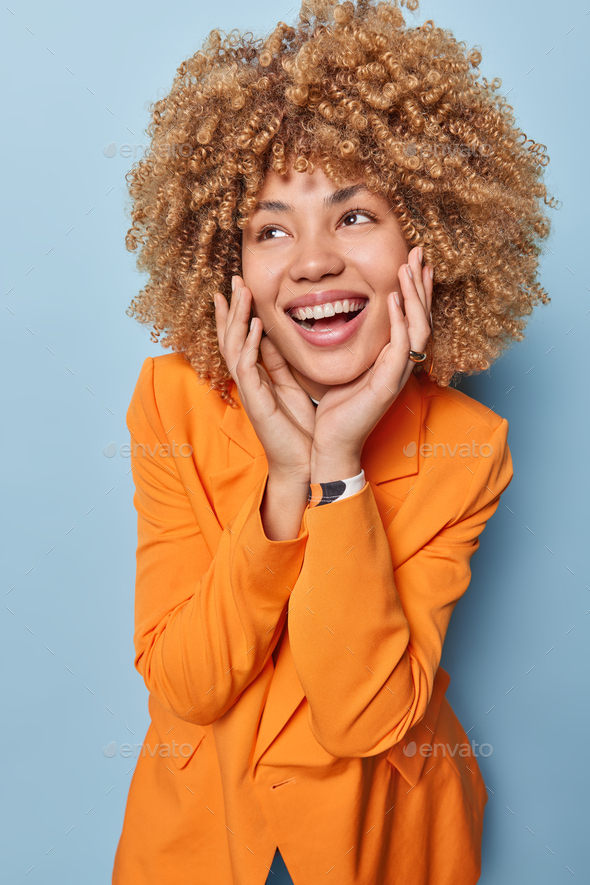 Happy dreamy woman dressed in formal orange jacket keeps hands on ...
