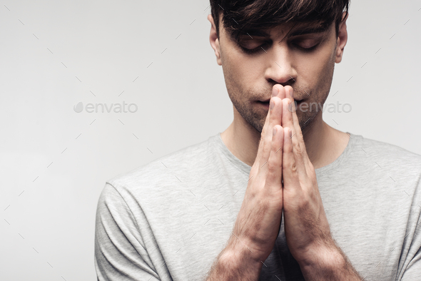 serious man with closed eyes showing praying gesture isolated on grey ...