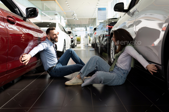 young couple fooling around in a car dealership sitting on the floor ...
