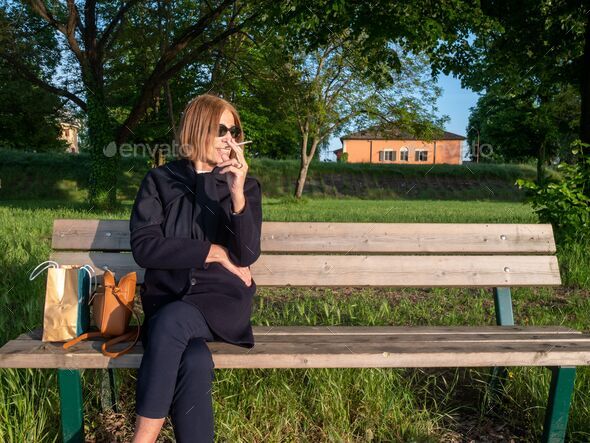 legs crossed senior woman smoking sitting on a park bench near a lake ...