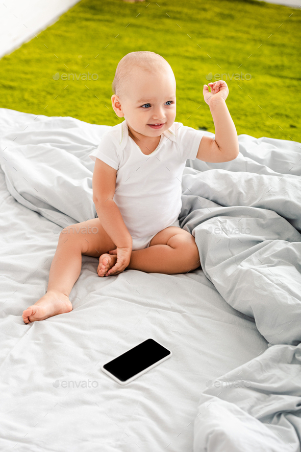 Adorable little child sitting on bed near digital device and raising ...