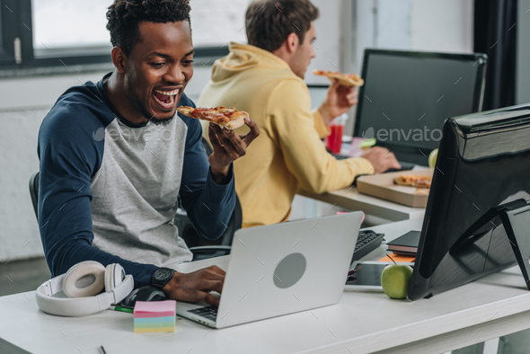 two multicultural programmers eating pizza while sitting near computers ...