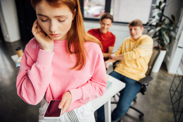 sad girl holding smartphone while classmates laughing at her in school ...