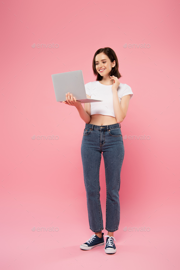 full length view of smiling pretty girl holding laptop isolated on pink ...