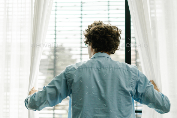Young man from behind, who opens the window curtains and looks out from ...