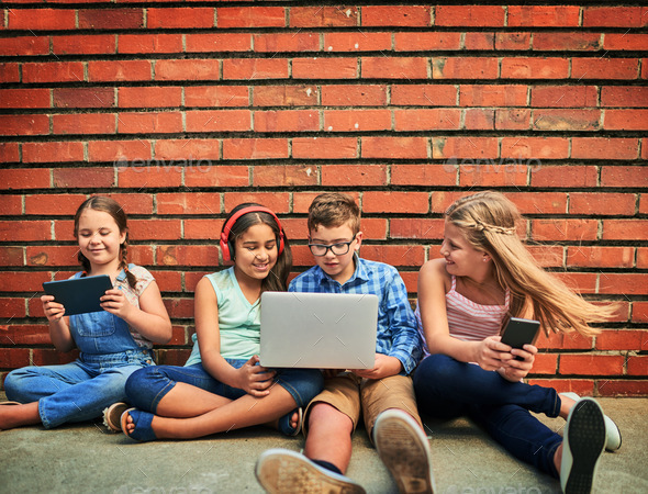 Shot of a group of young children using digital devices against a brick ...