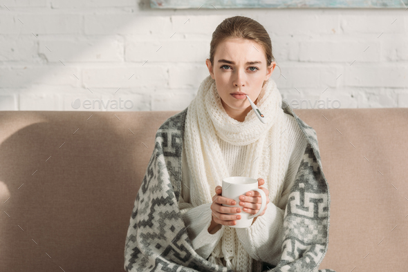 sick girl looking at camera while measuring temperature and holding cup ...