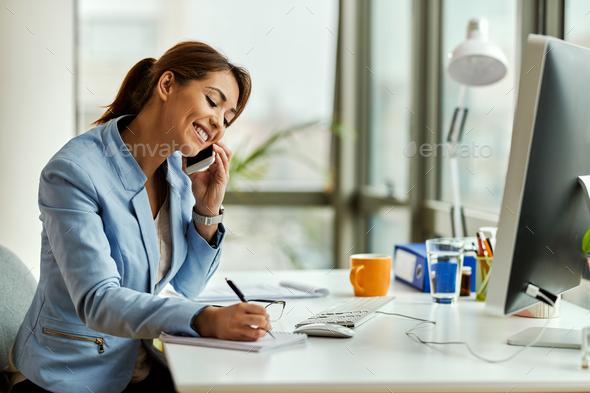 Happy businesswoman writing notes while talking on the phone in the ...
