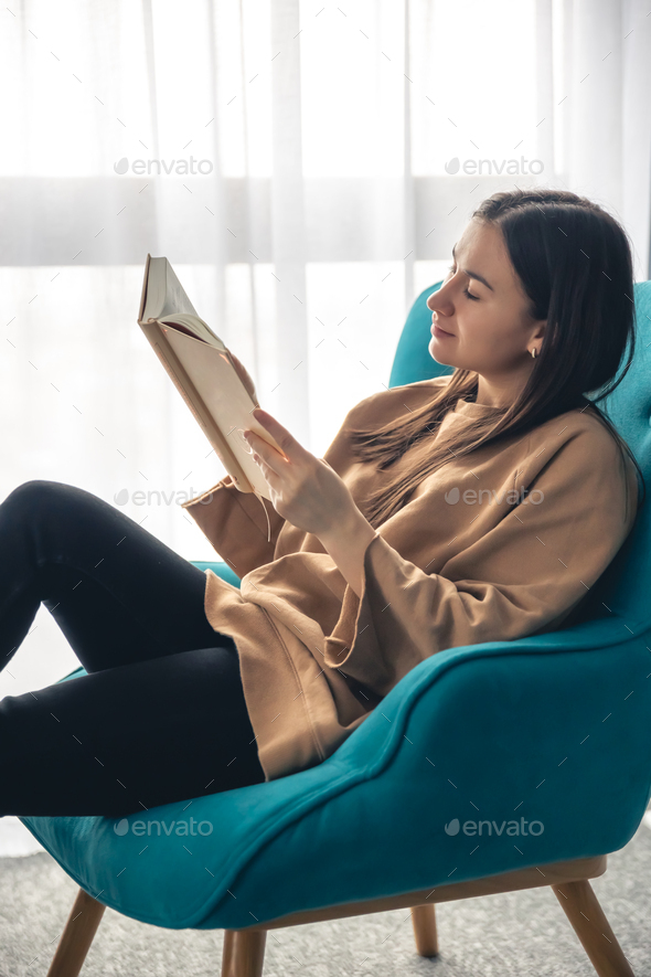 A young woman is reading a book while sitting on an armchair by the ...