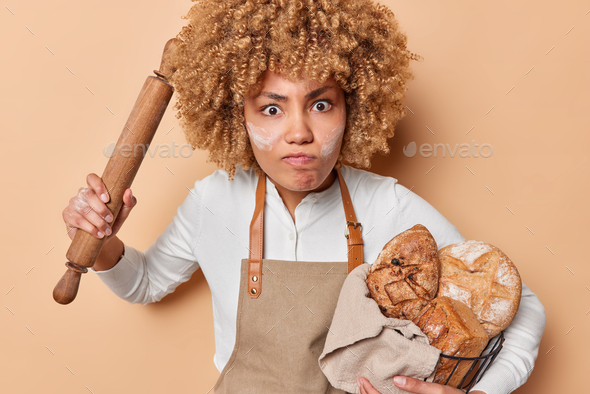 Angry curly haired female baker holds rolling pin and basket of bread ...