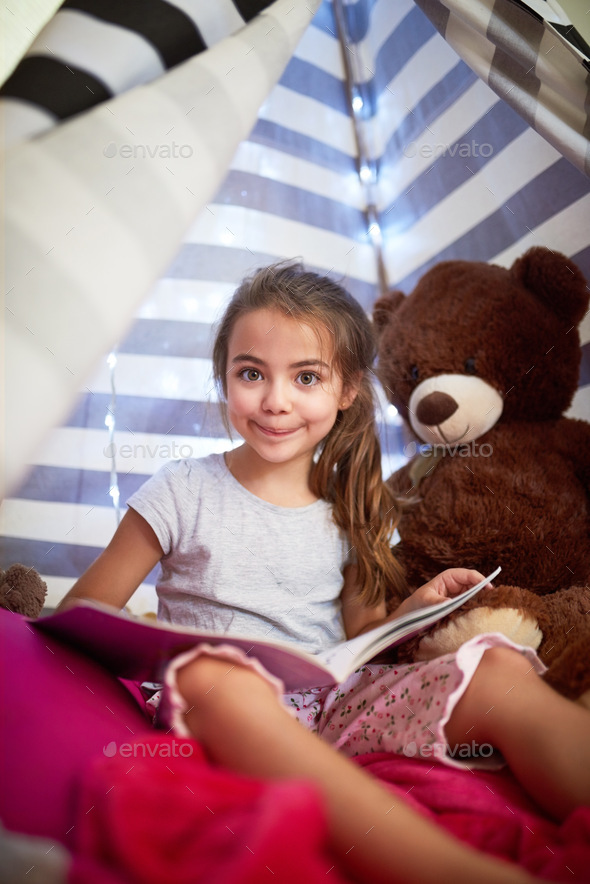 Kids will be kids. Portrait of a little girl reading a book with her teddybear in a tent at home ...