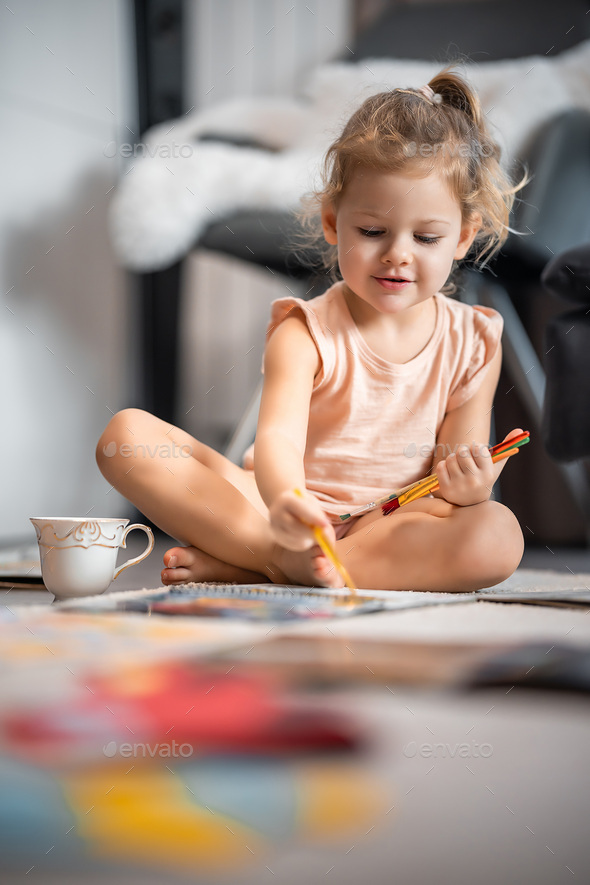 Little girl sits on the floor at home and draws with paints and brushes ...