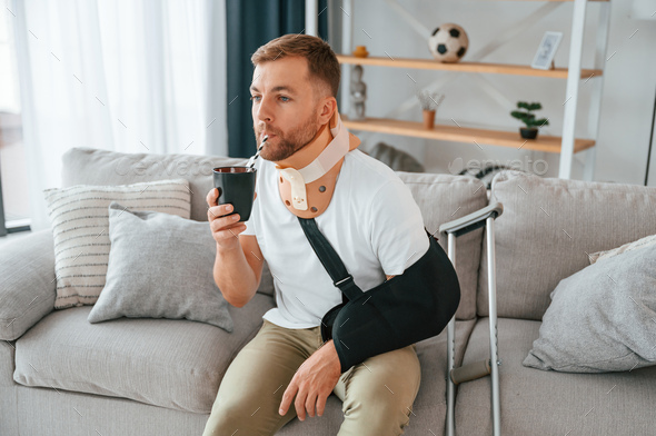 Drinking from cup by using swizzle stick. Man with crutches is at home ...