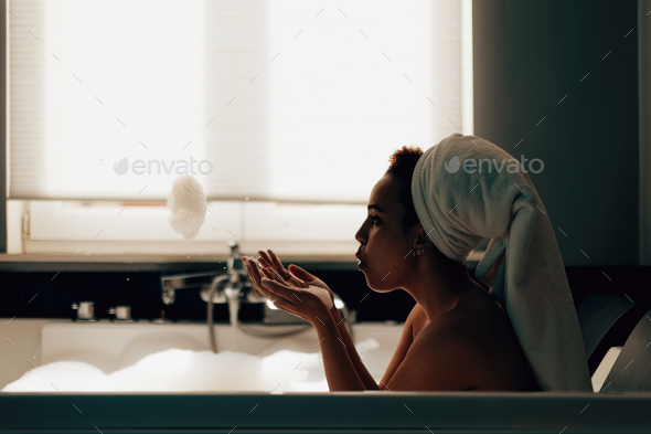 young lady in backlight taking a foam bath in tub and blowing on the ...