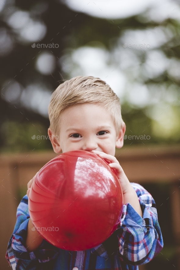 Vertical shot of a happy child blowing up a red balloon with a blurred ...