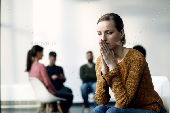 Young sad woman thinking while sitting apart from participants of group ...