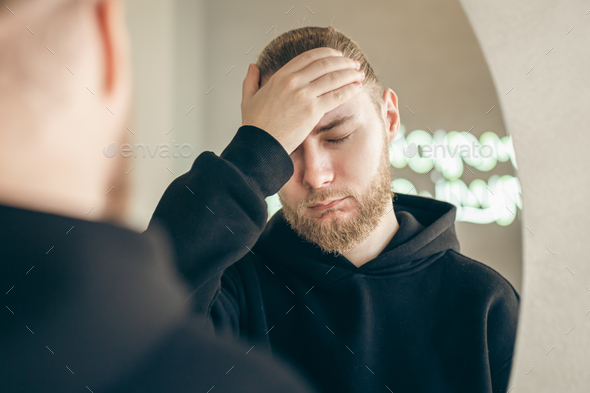 Sad young man with a beard in front of a mirror, mental health concept ...