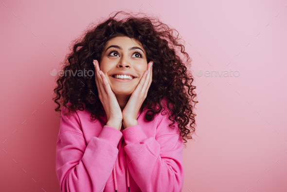 happy bi-racial girl touching face and looking up on pink background ...