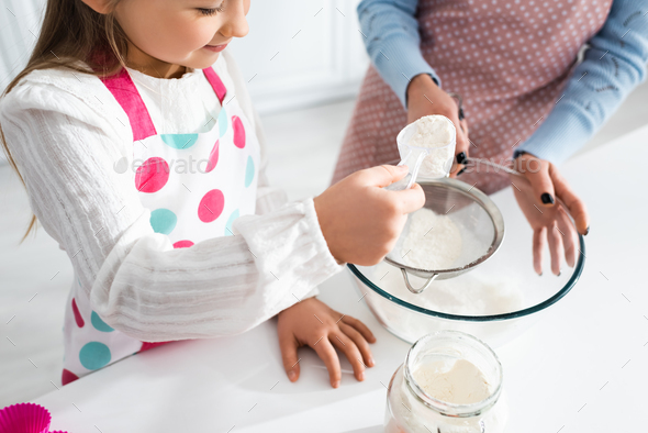 cropped view of mother holding sieve and smiling daughter sifting flour ...
