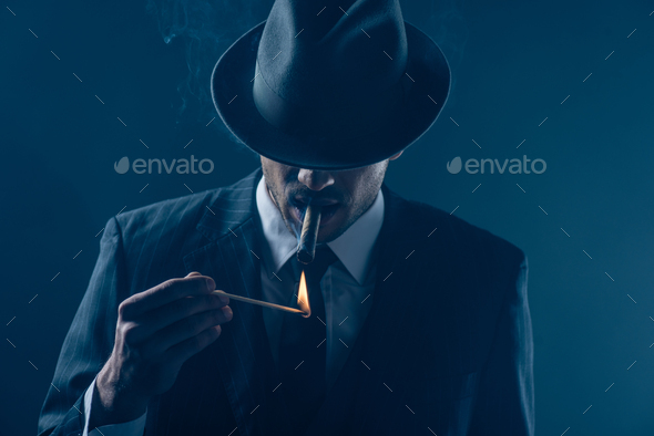 Mafioso with covered eyes with felt hat lighting cigar on dark blue ...