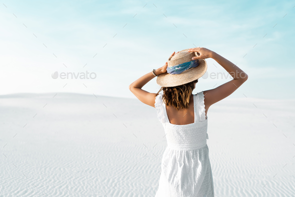back view of beautiful girl in white dress and straw hat on sandy beach ...