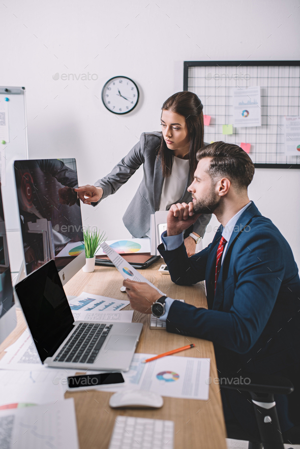 Data analyst pointing at computer monitor near colleague holding paper with charts at table ...