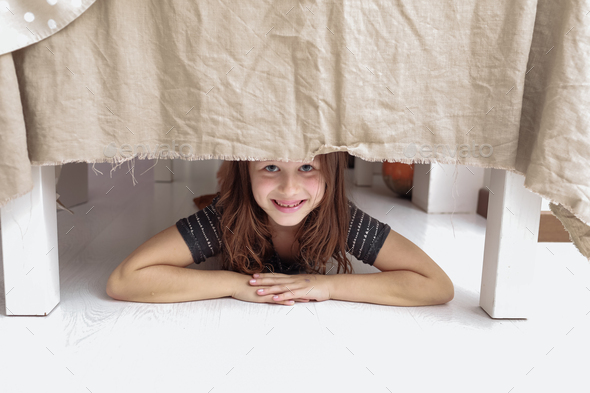 Portrait of cute small girl lying on floor under table indoors at home ...